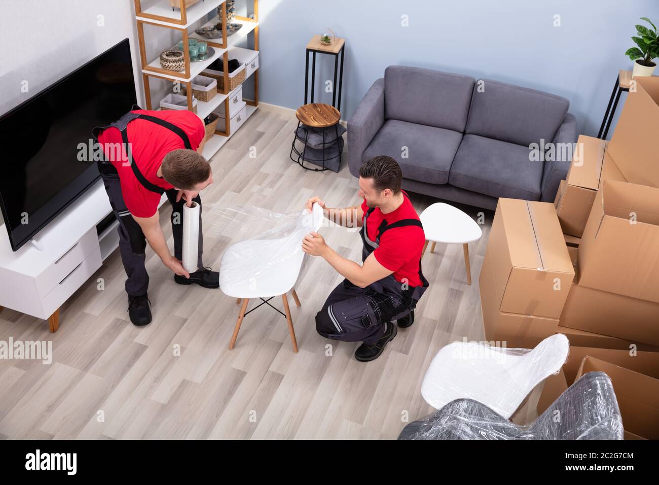 Two Young Male Movers Packing White Chair With Plastic Wrap In Living ...