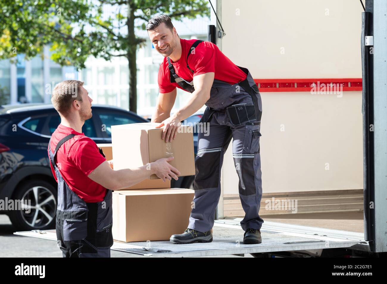 Two Young Male Movers In Uniform Carrying Cardboard Boxes From Truck ...