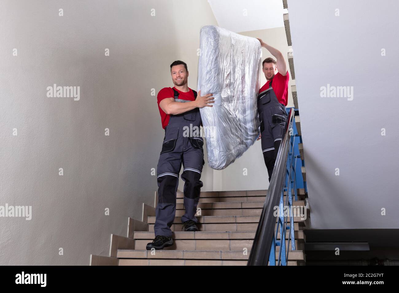Two Young Male Movers In Uniform Carrying The Wrapped Mattress While ...