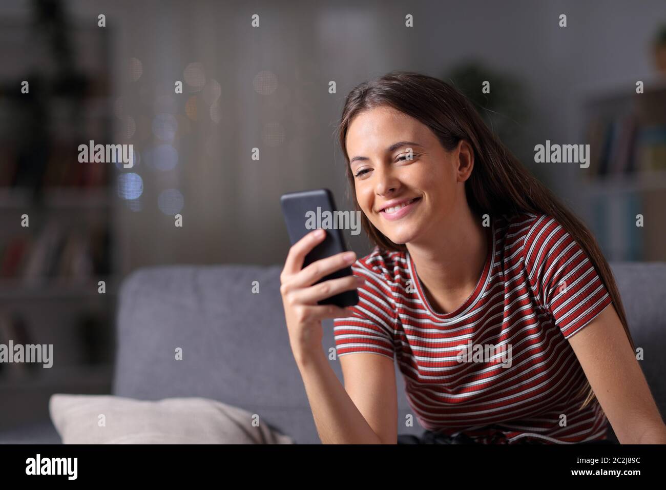 Happy teen in the night using mobile phone sitting on a couch in the ...