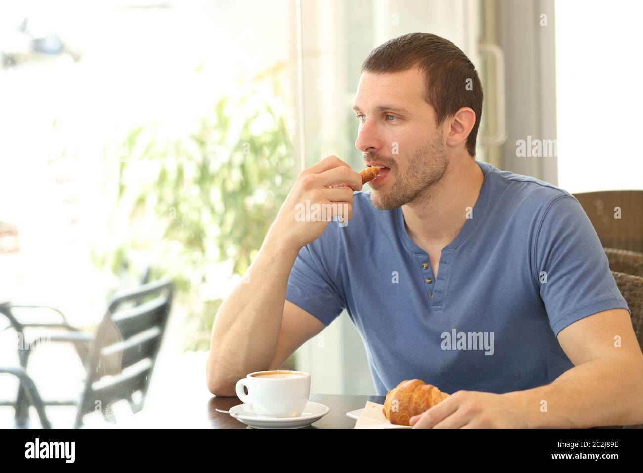 Relaxed man eating a croissant for breakfast sitting in a coffee shop ...