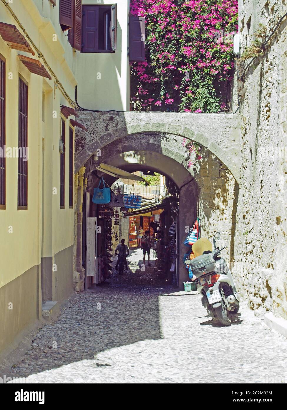 people walk past shops and old arches in a narrow street in rhodes town ...