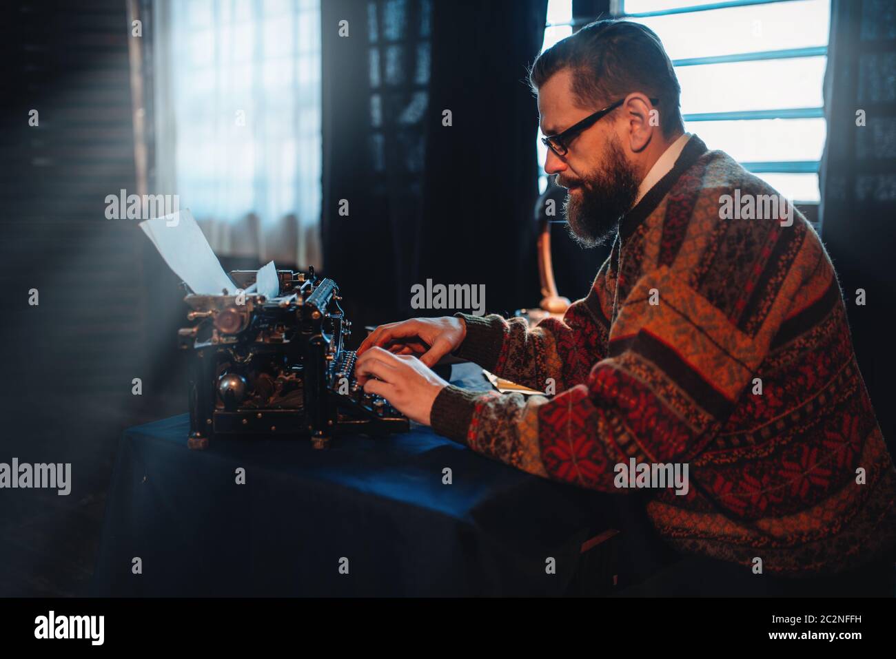 Bearded writer in glasses typing on retro typewriter against window ...