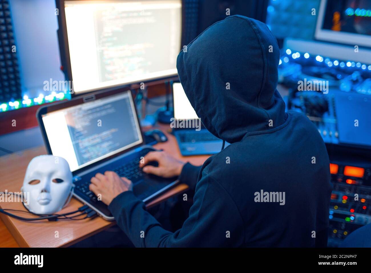 Hacker in mask and hood sitting at his workplace with laptop and PC ...