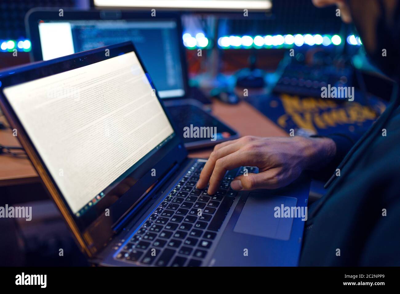 Hacker in hood shows thumbs up at his workplace with laptop and PC ...