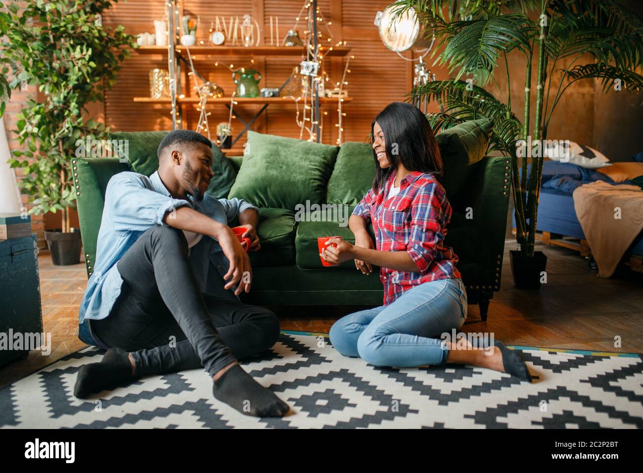 Black couple sitting on the floor and drinks coffee against a couch at ...
