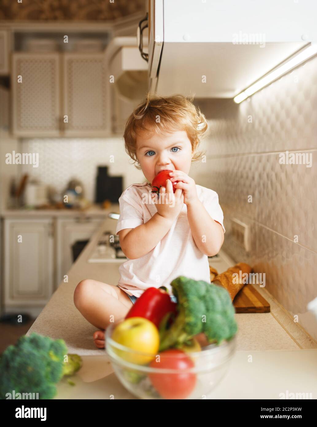 Little girl sitting on the table and eats tomato. Female baby tasting ...