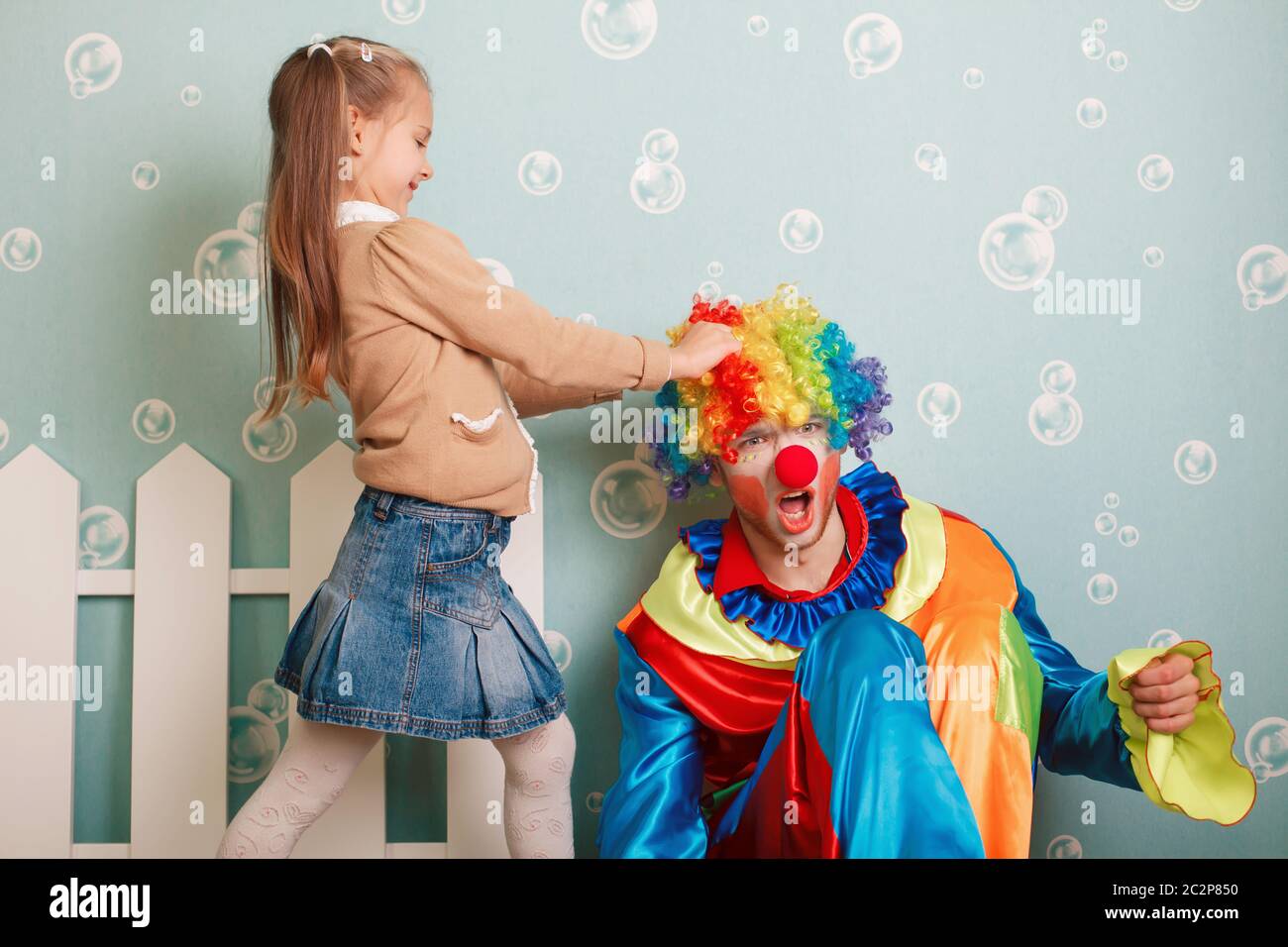 Little girl pulls clown hair. White fence decoration on the background ...