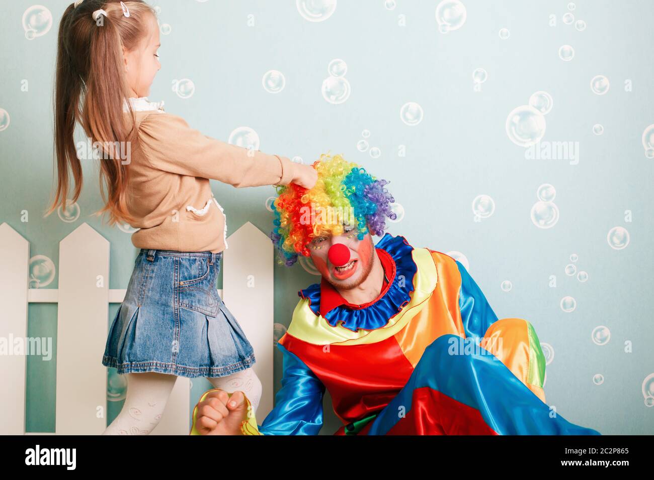 Little girl pulls clown hair. White fence decoration on the background ...