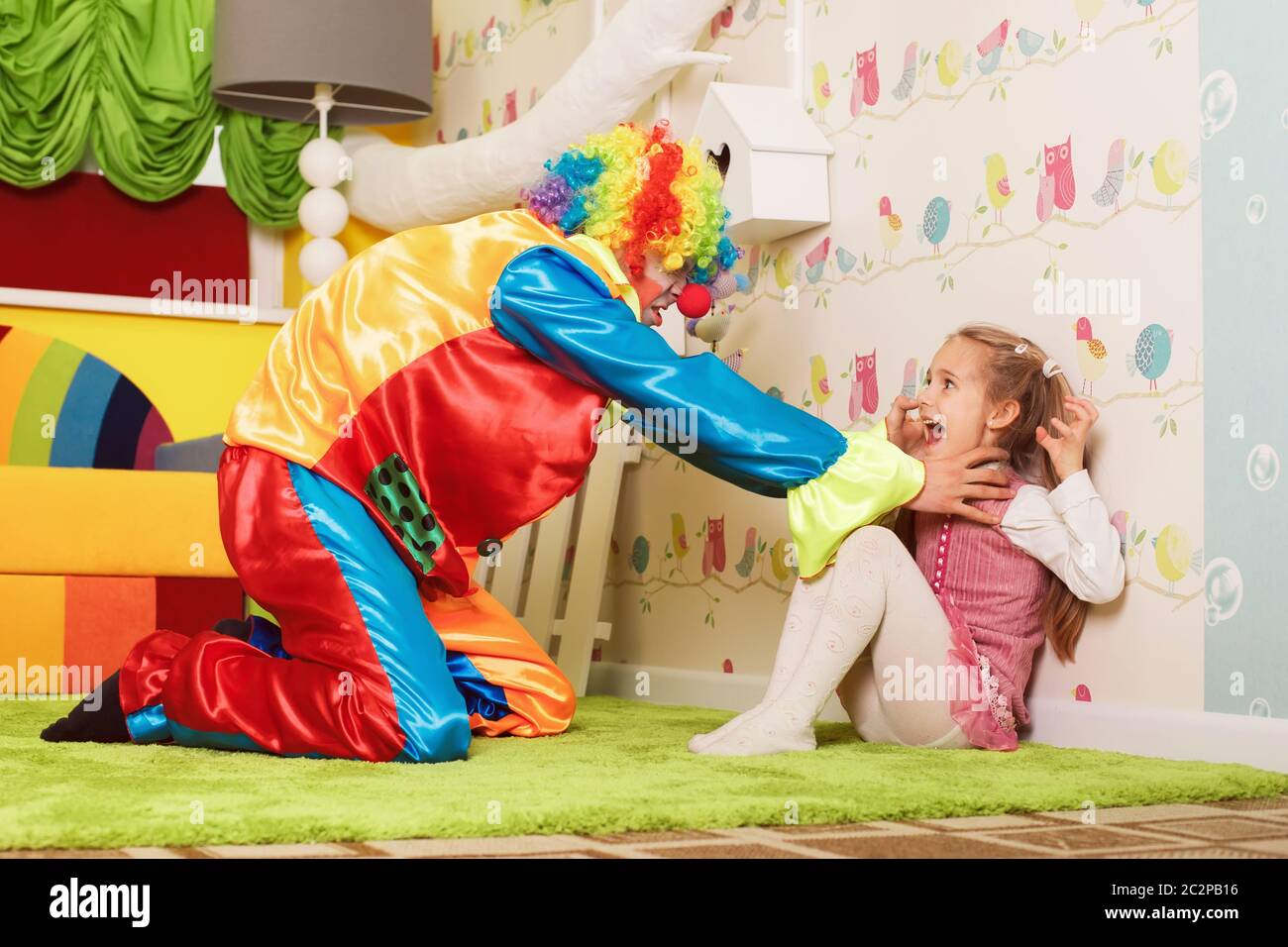 Frightened girl and a clown in colorful costume playing on the green ...