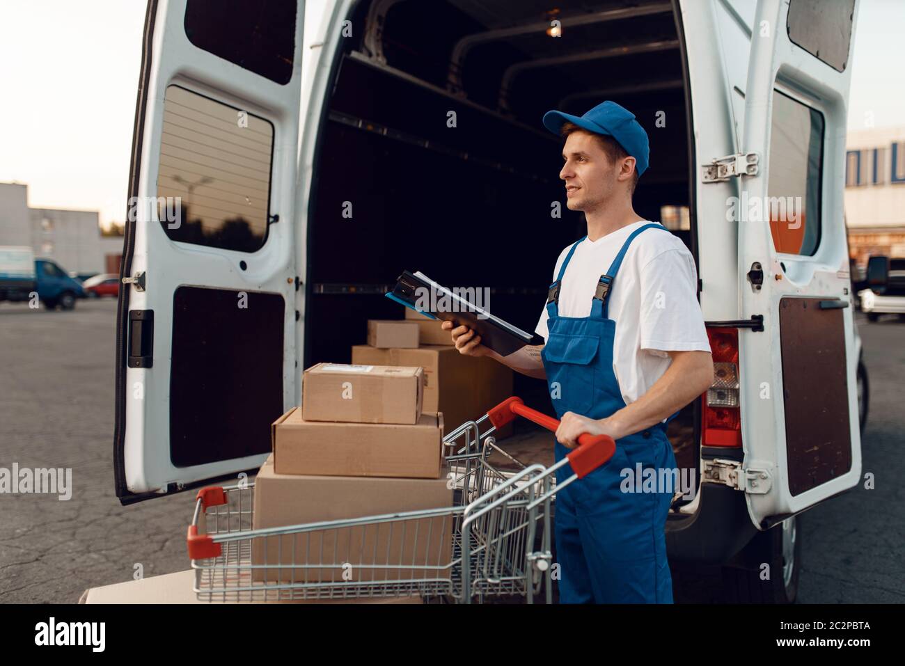 Deliveryman in uniform holds cart with boxes at the car with parcels ...