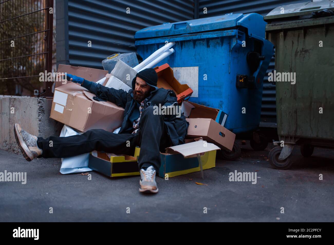 Dirty drunk beggar lies in garbage at the trashcan on city street ...