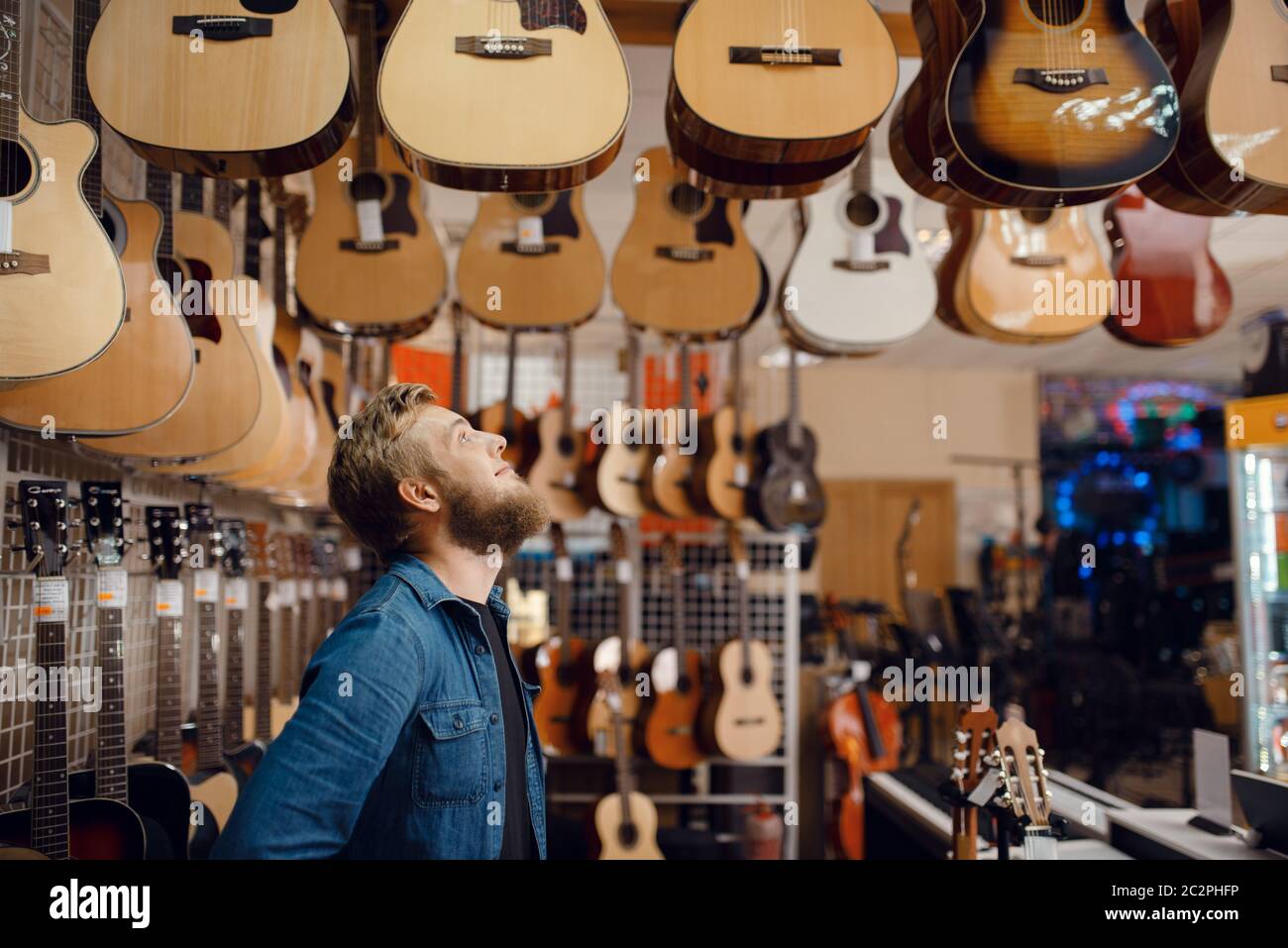 Bearded young guy choosing acoustic guitar in music store. Assortment ...