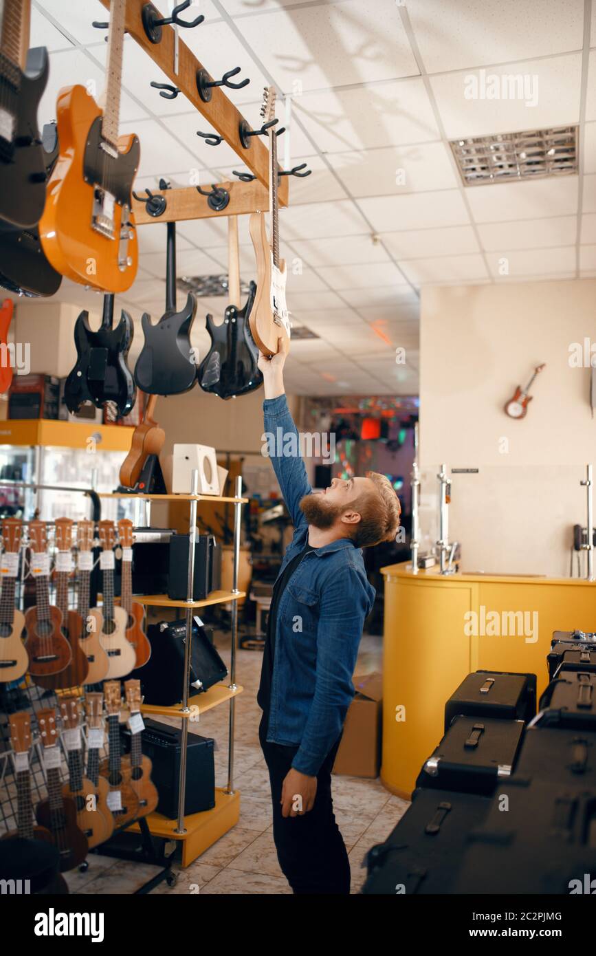 Bearded young man choosing acoustic guitar in music store. Assortment ...