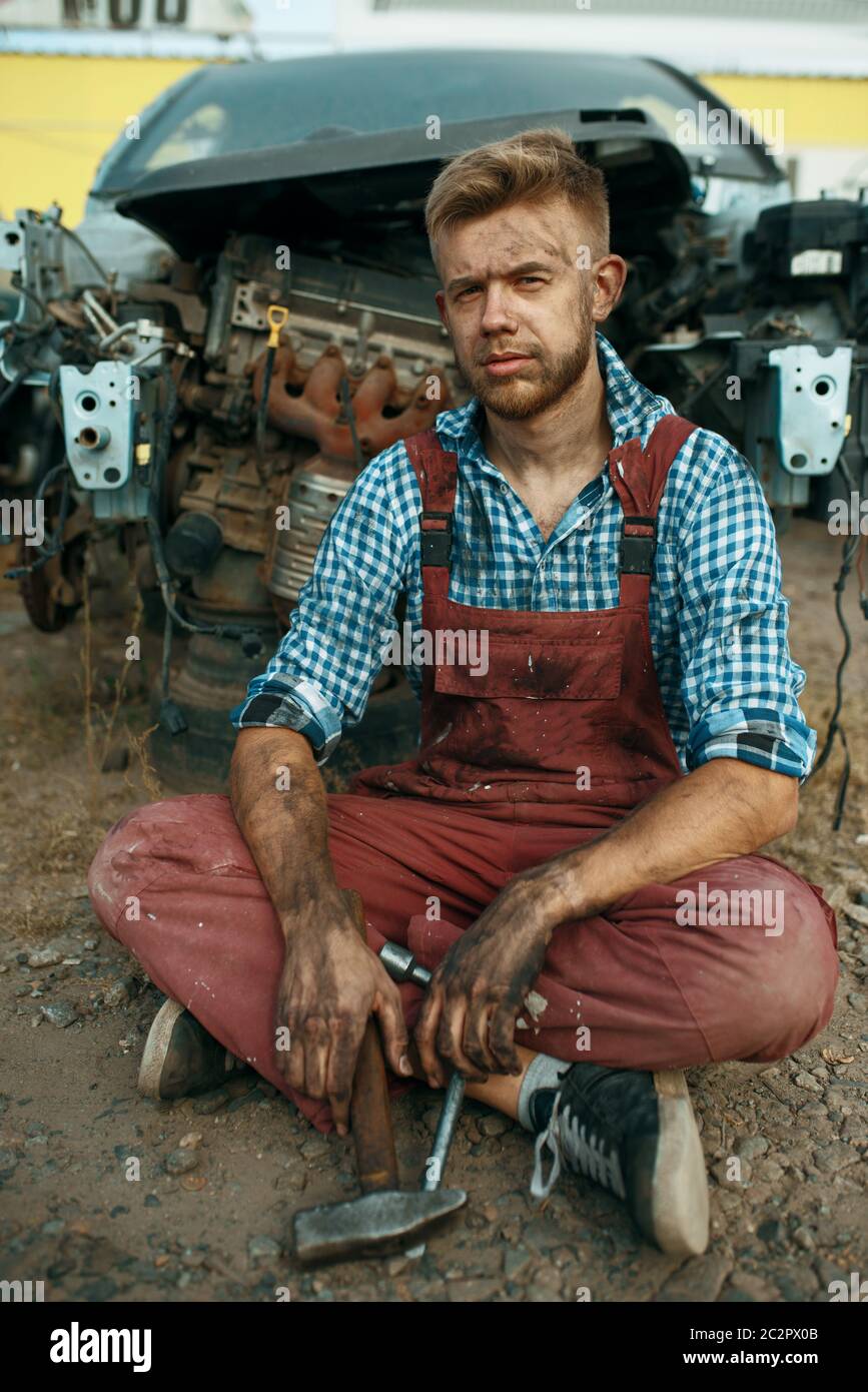 Male repairman sitting on the ground on car junkyard. Auto scrap ...