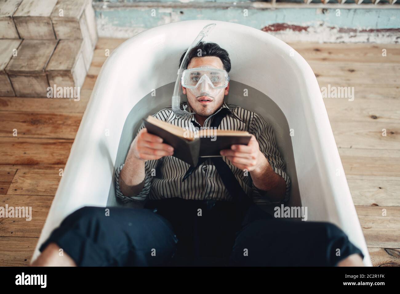Funny businessman in flippers and mask lies in bathtub and reads a book ...
