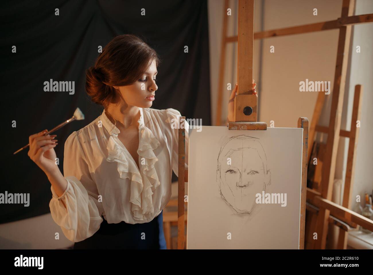 Cute female painter with brush standing against easel in studio ...