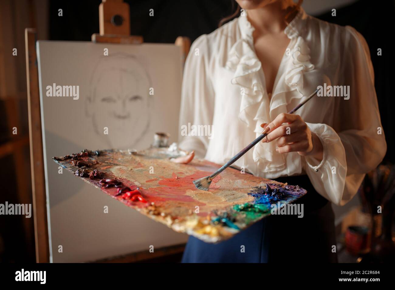 Female painter with color palette and brush standing against easel in ...