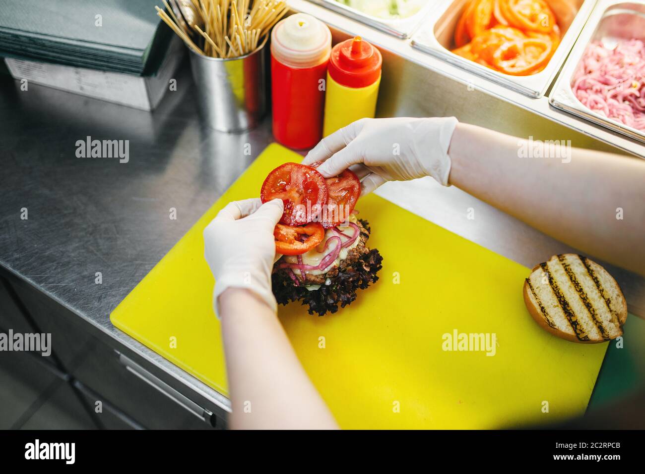Male cook puts tomatoes in to the burger, fast food cooking. Hamburger ...