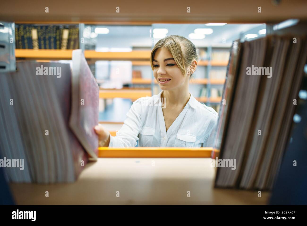 Female student choosing book at the bookshelf in university library ...