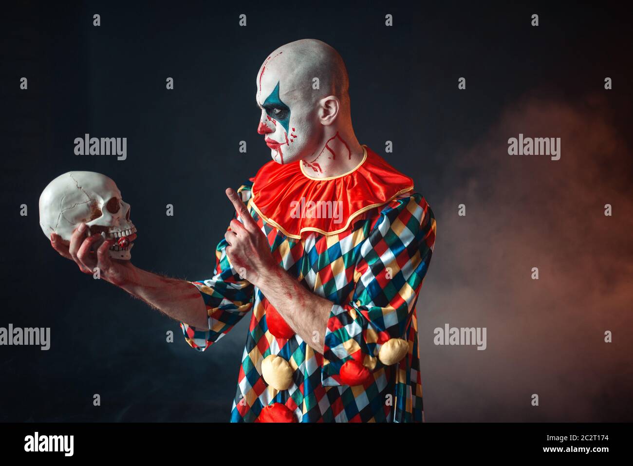 Mad bloody clown holds human skull in red wig, horror. Man with makeup ...