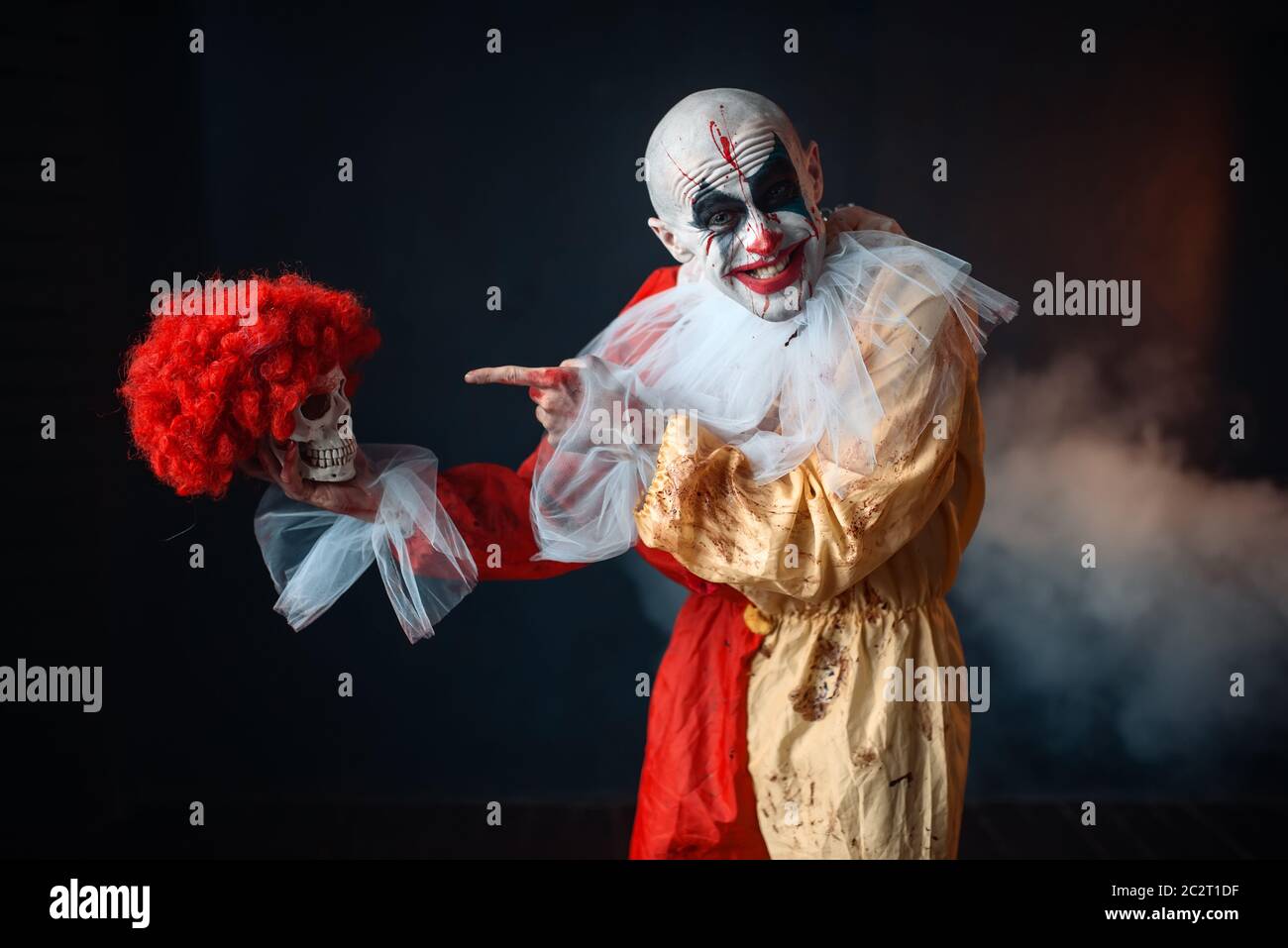 Mad bloody clown holds human skull in red wig, horror. Man with makeup ...