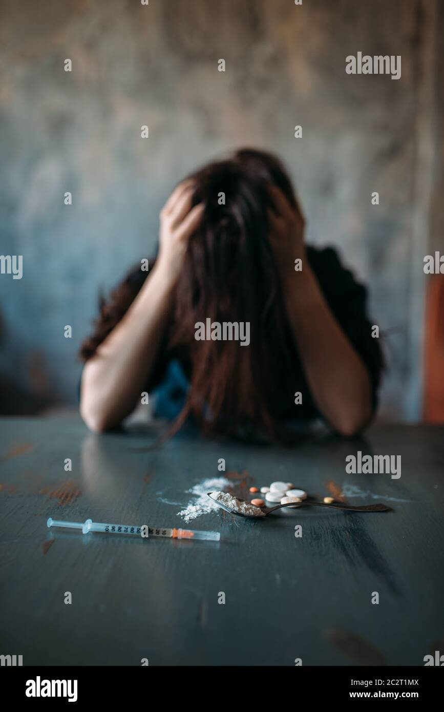 Female junkie sitting at the table with drugs and syringe, grunge room ...