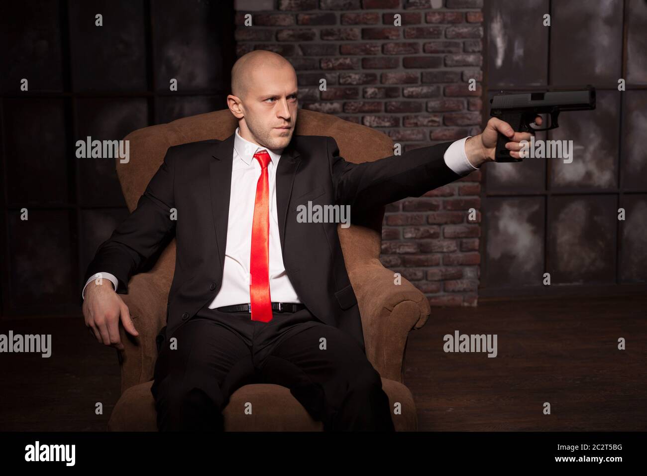 Bald killer in suit and red tie sitting in chair and aims a pistol ...