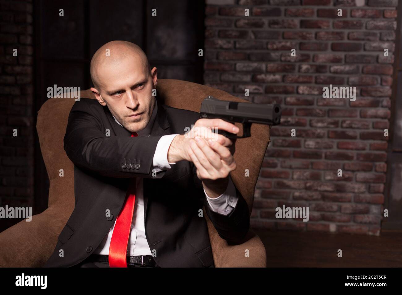 Bald killer in suit and red tie sitting in chair and aims a pistol ...
