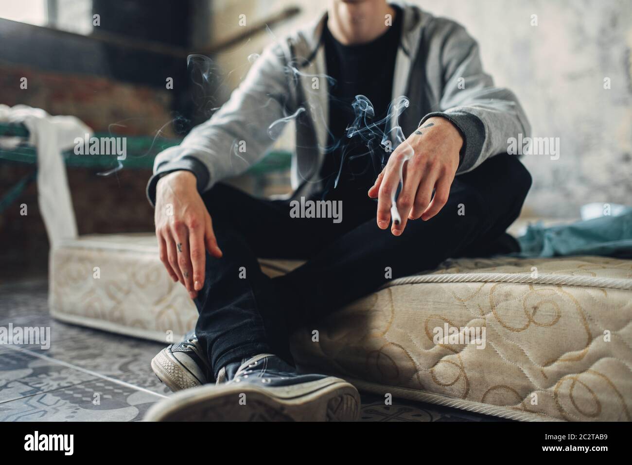 Male junkie sitting on the mattress and smoke cigarette, grunge room ...