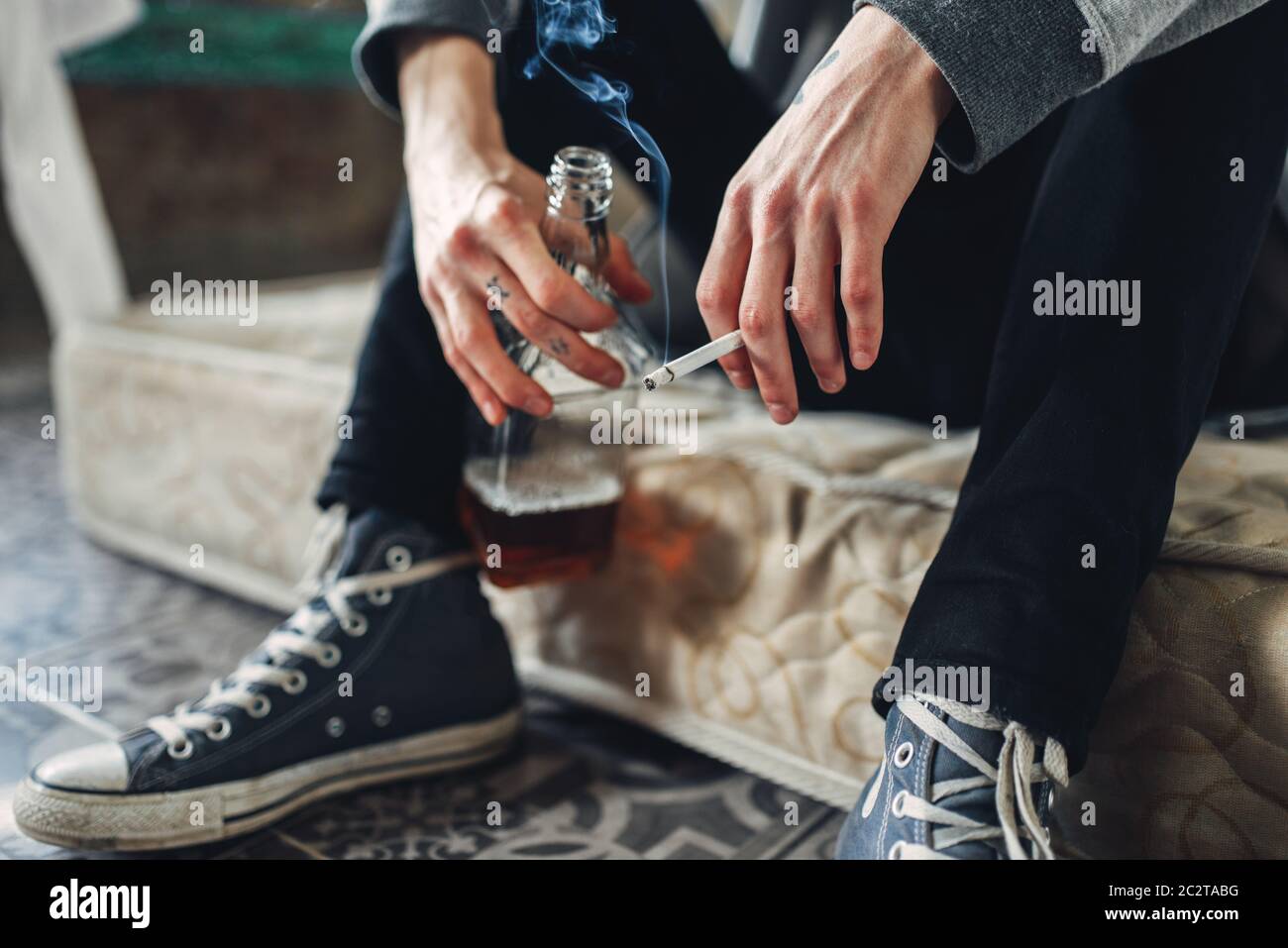Addicted male person sitting on the mattress with cigarette and bottle ...