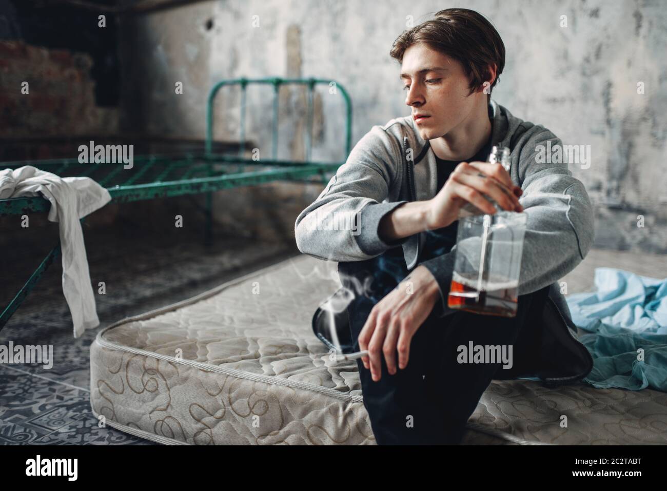 Male druggy sitting on the mattress, smoke cigarette and drinks alcohol ...