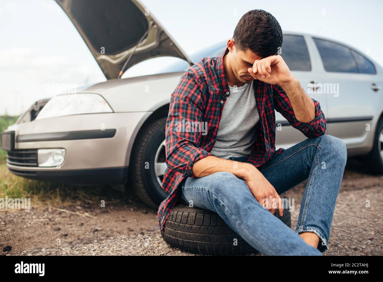 Tired man sitting on tire, broken car with open hood on background ...