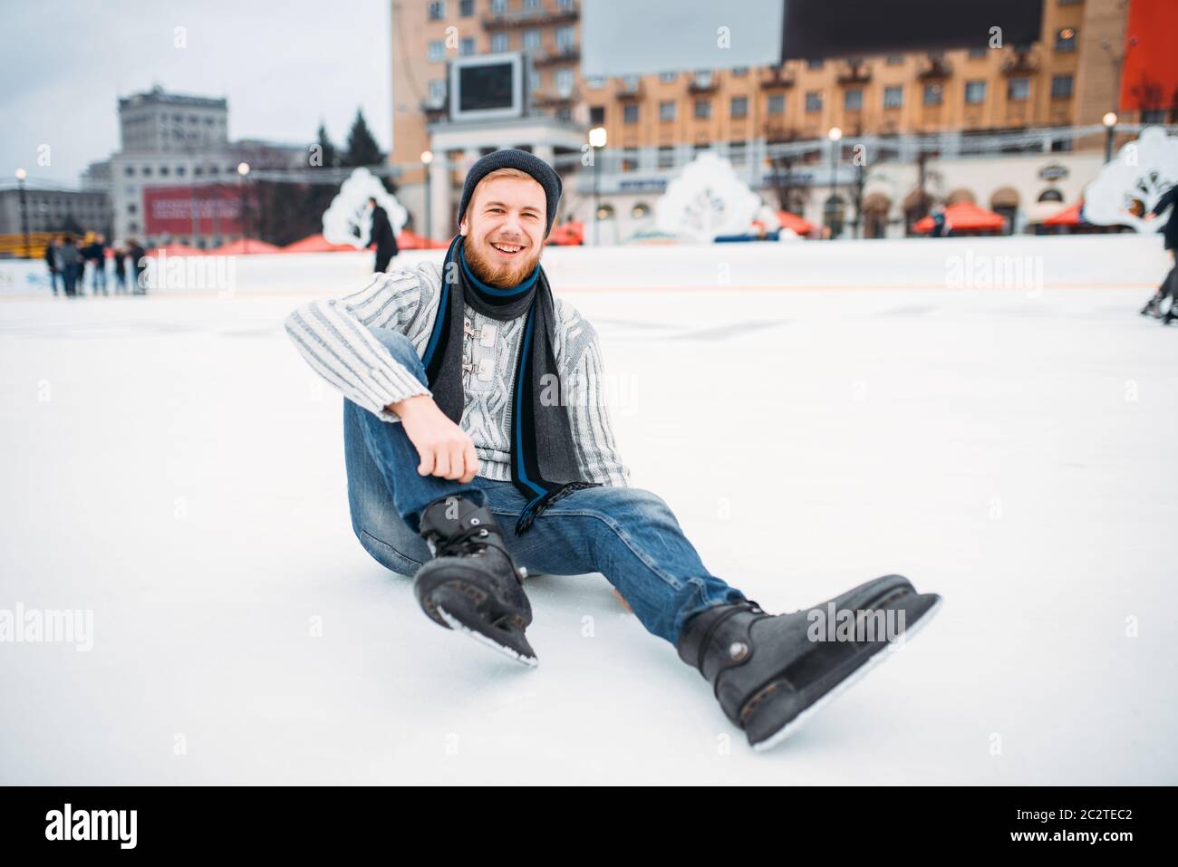 Young smiling man in skates sitting on ice, skating rink. Winter ice ...