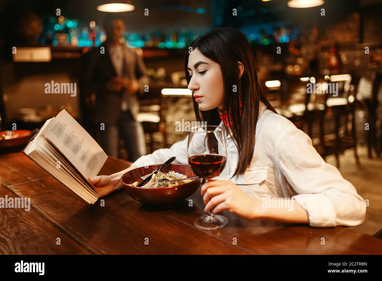 Young woman drinks red wine and reading a book at wooden bar counter ...