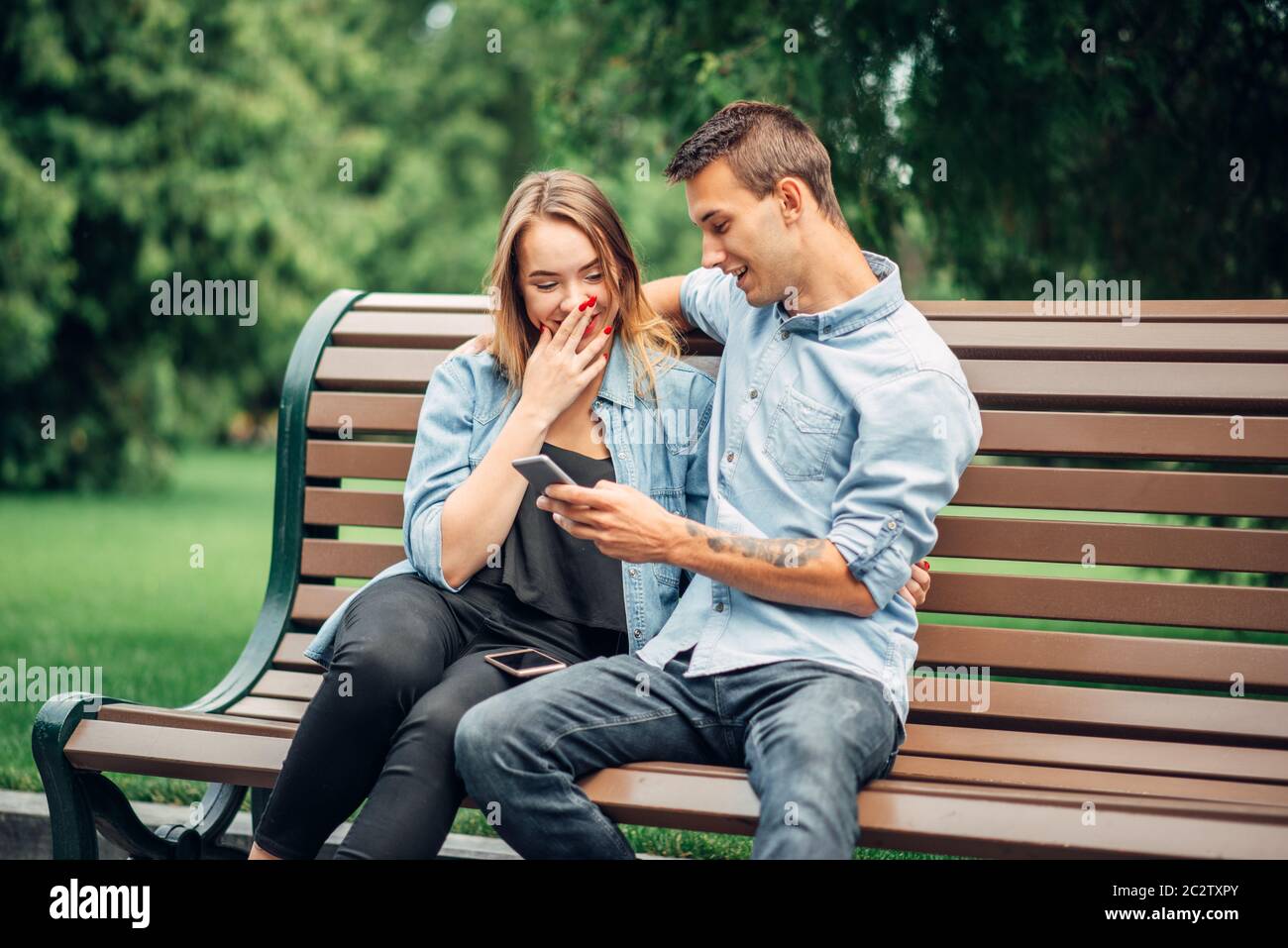 Phone addiction, smiling couple using gadget on the bench in summer ...