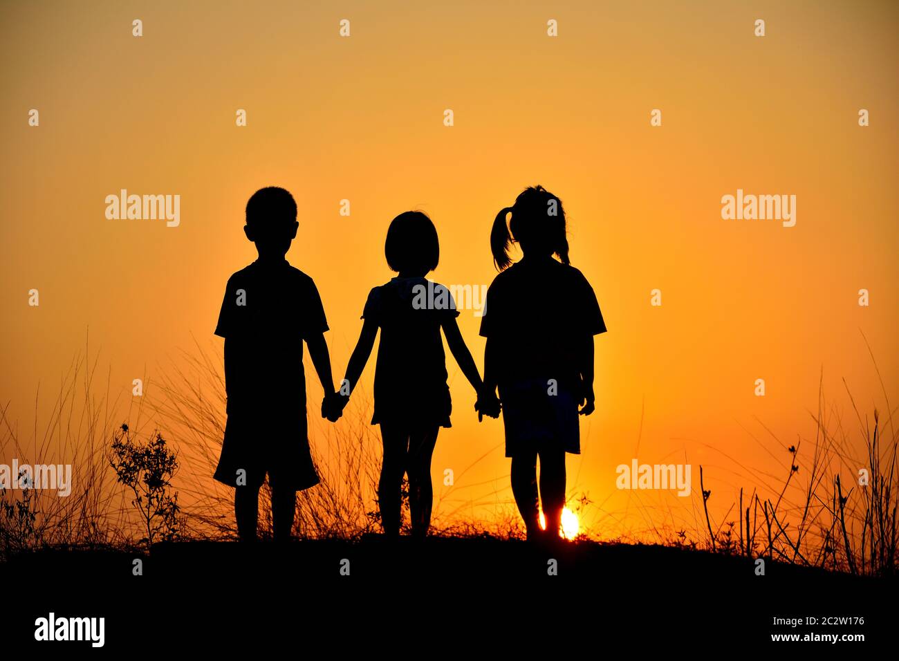 Black silhouette of three children standing together. There is a sky at ...