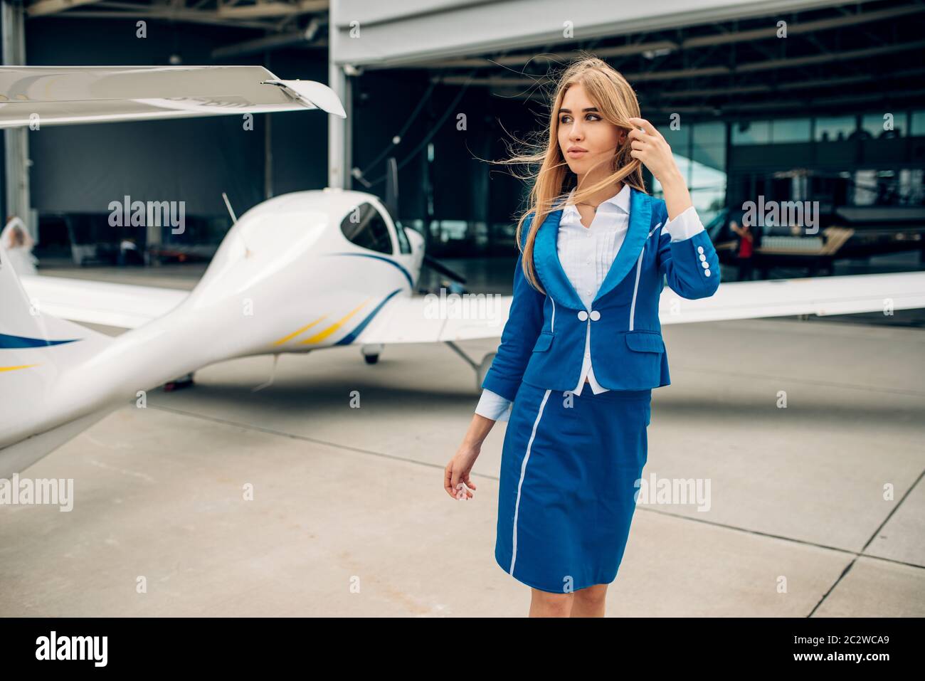 Attractive stewardess in uniform poses against small airplane in hangar ...