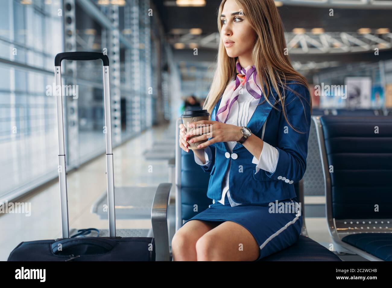 Stewardess with coffee and suitcase sitting on seat in waiting area in ...