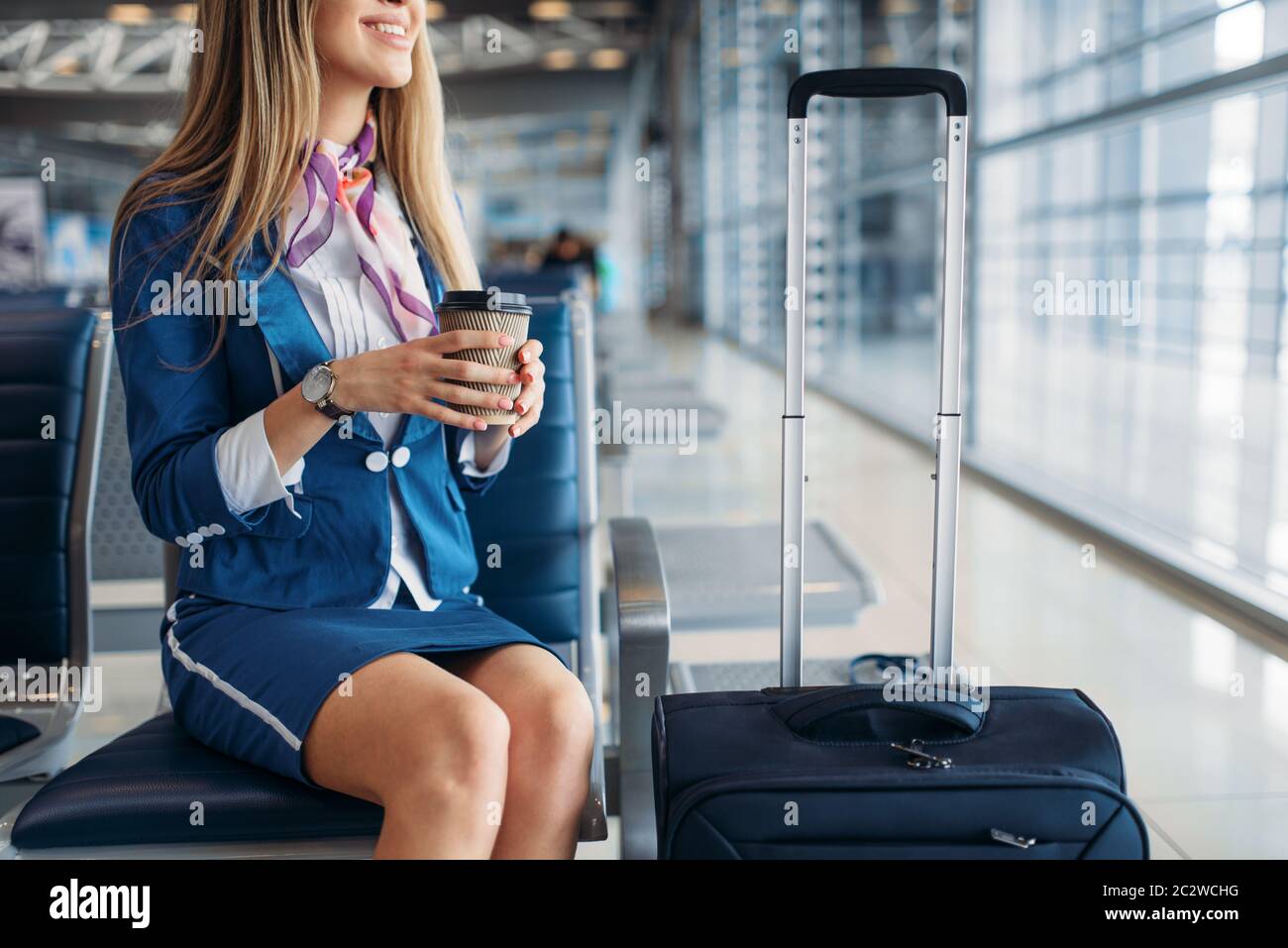 Stewardess with coffee and suitcase sitting on seat in waiting area in ...