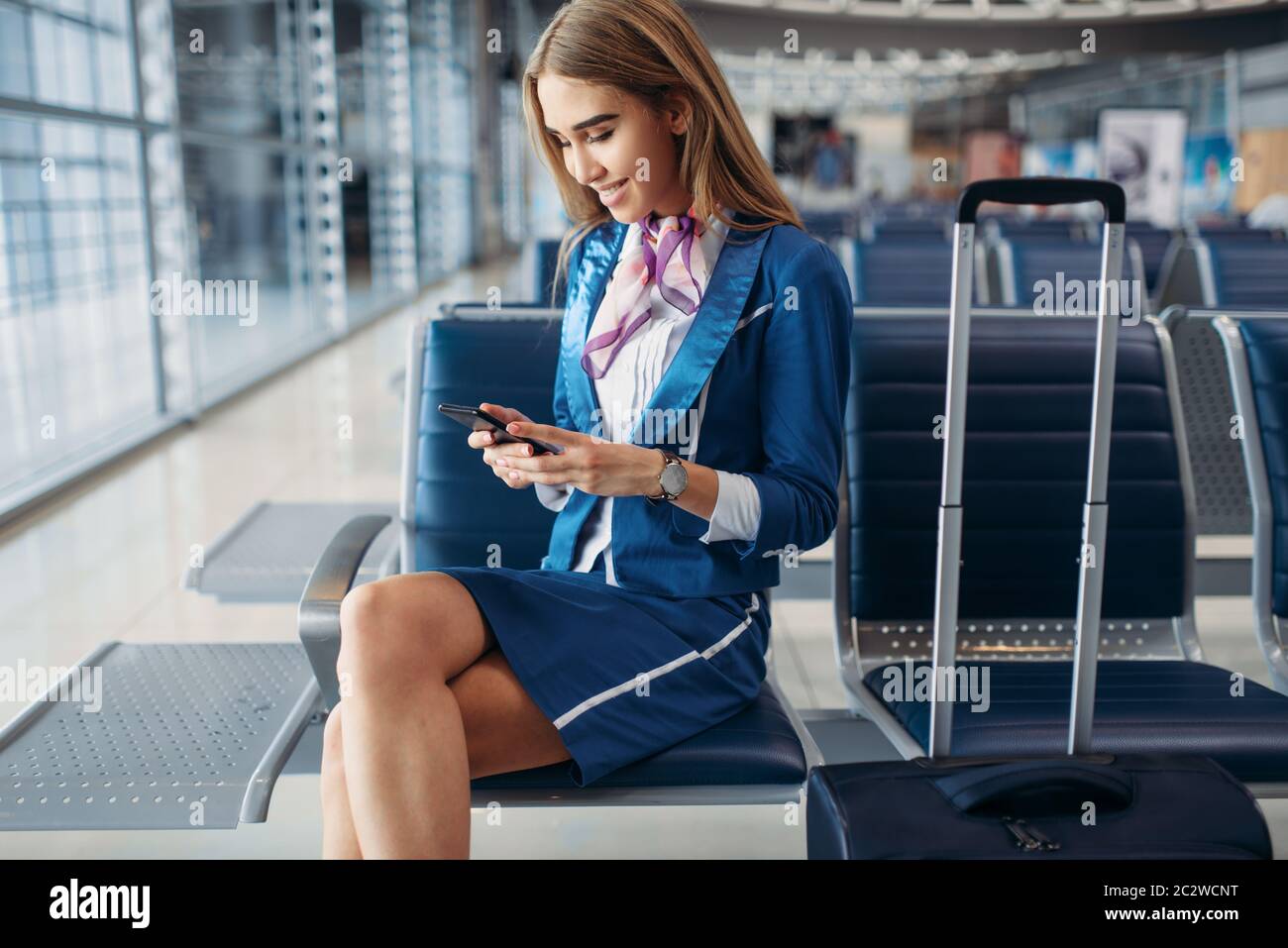 Stewardess with suitcase sitting on seat in waiting area in airport and ...