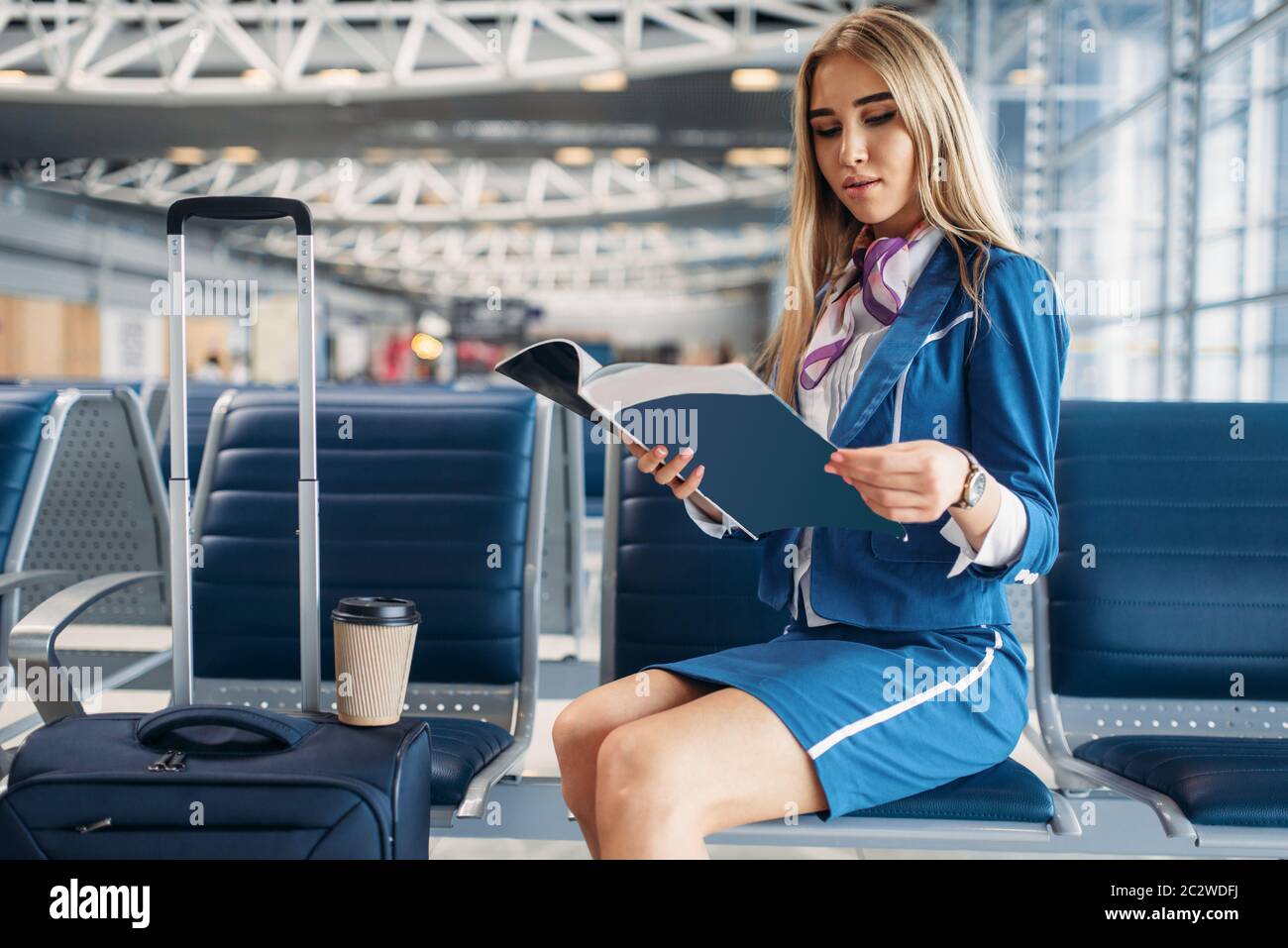 Stewardess with suitcase sitting on seat in waiting area in airport and ...