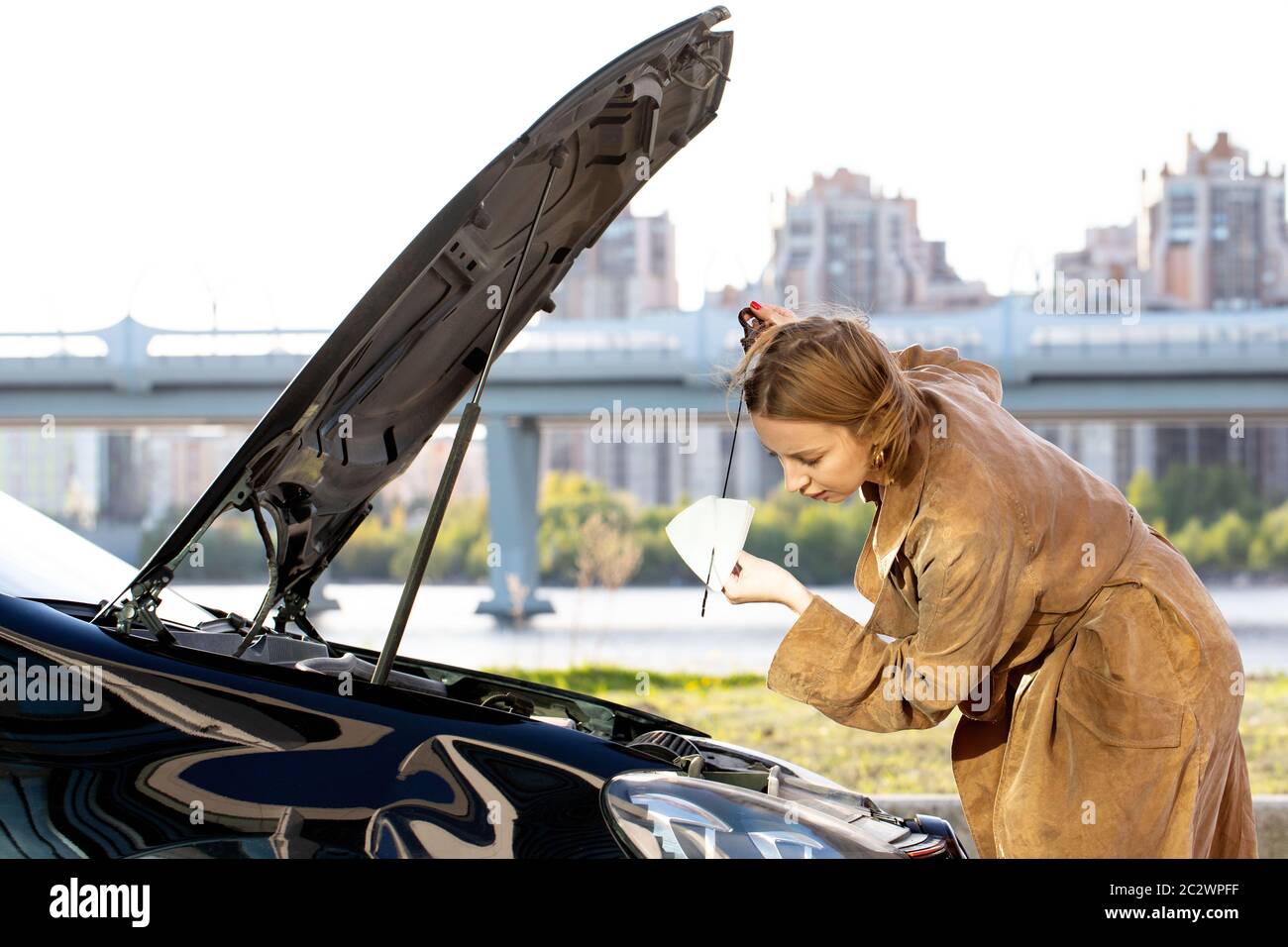 Confident caucasian woman driver checking oil level in car engine dipstick, cityscape on ...