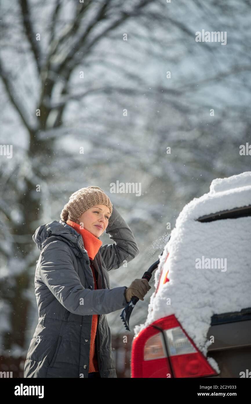 Pretty, young woman cleaning her car from snow after heavy snowstorm ...
