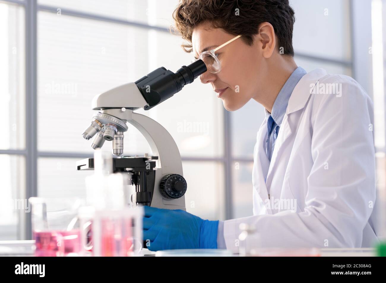 Focused microbiologist in glasses sitting at desk and using microscope ...