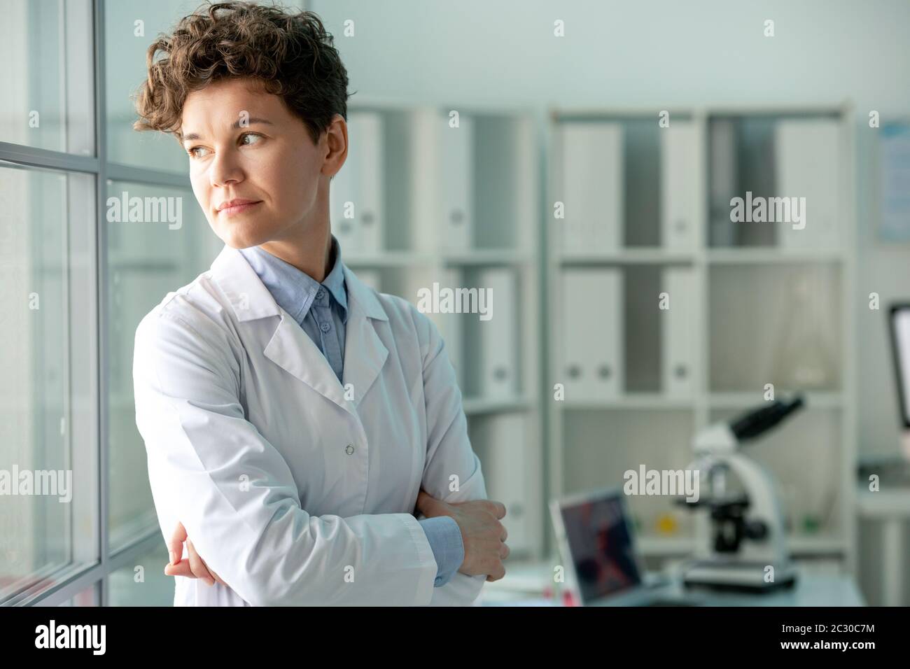 Pensive middle-aged female researcher in lab coat standing with crossed ...