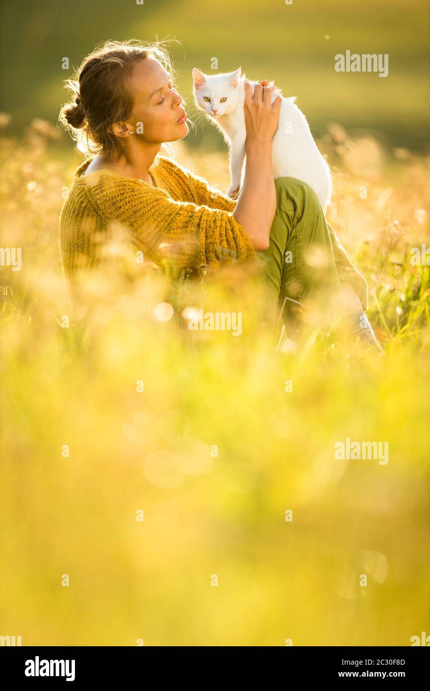 Pretty, young woman with her cat pet sitting in grass on lovely meadow ...