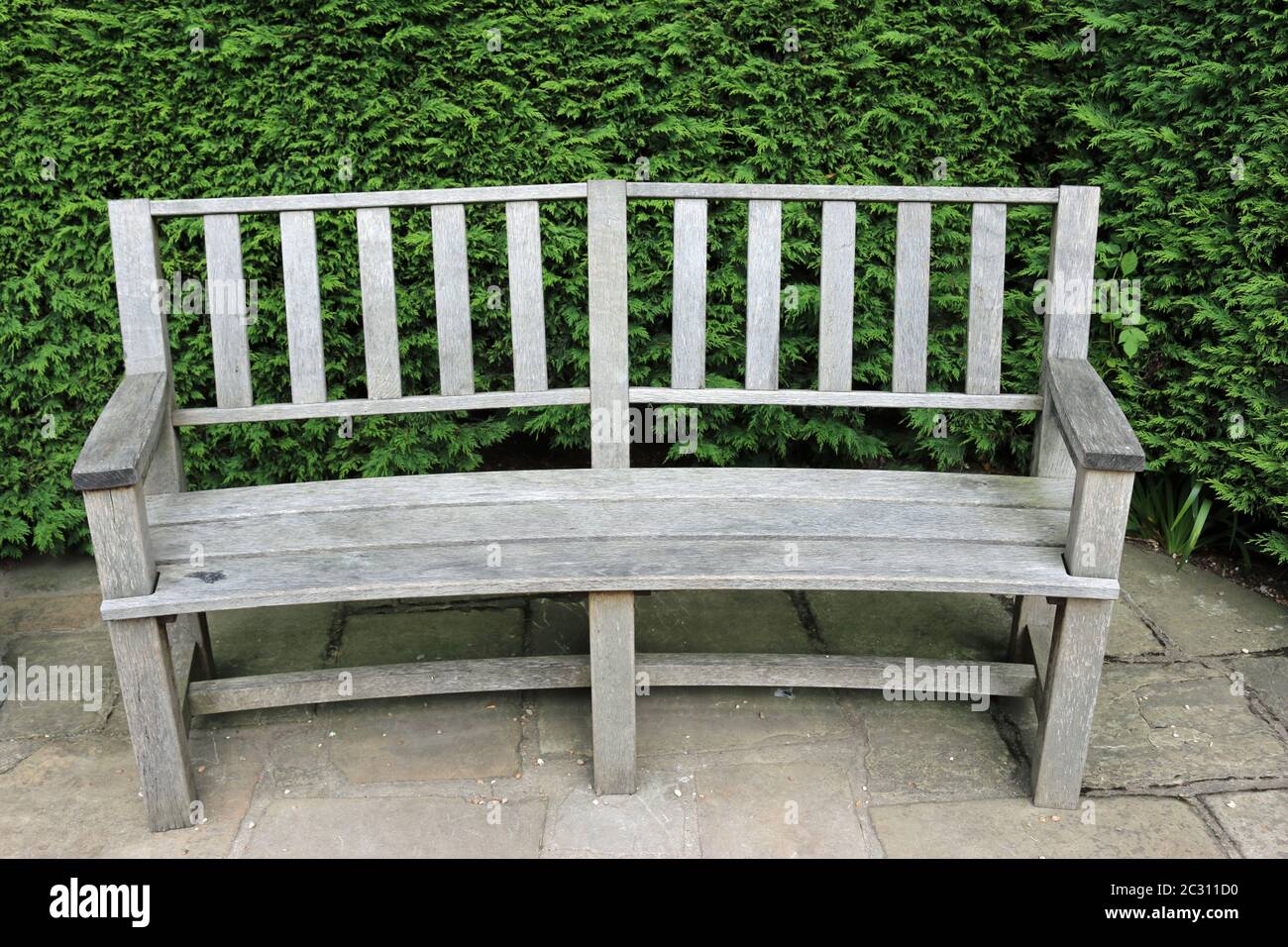 Curved wooden park seat on stone paving slabs with conifers in the ...