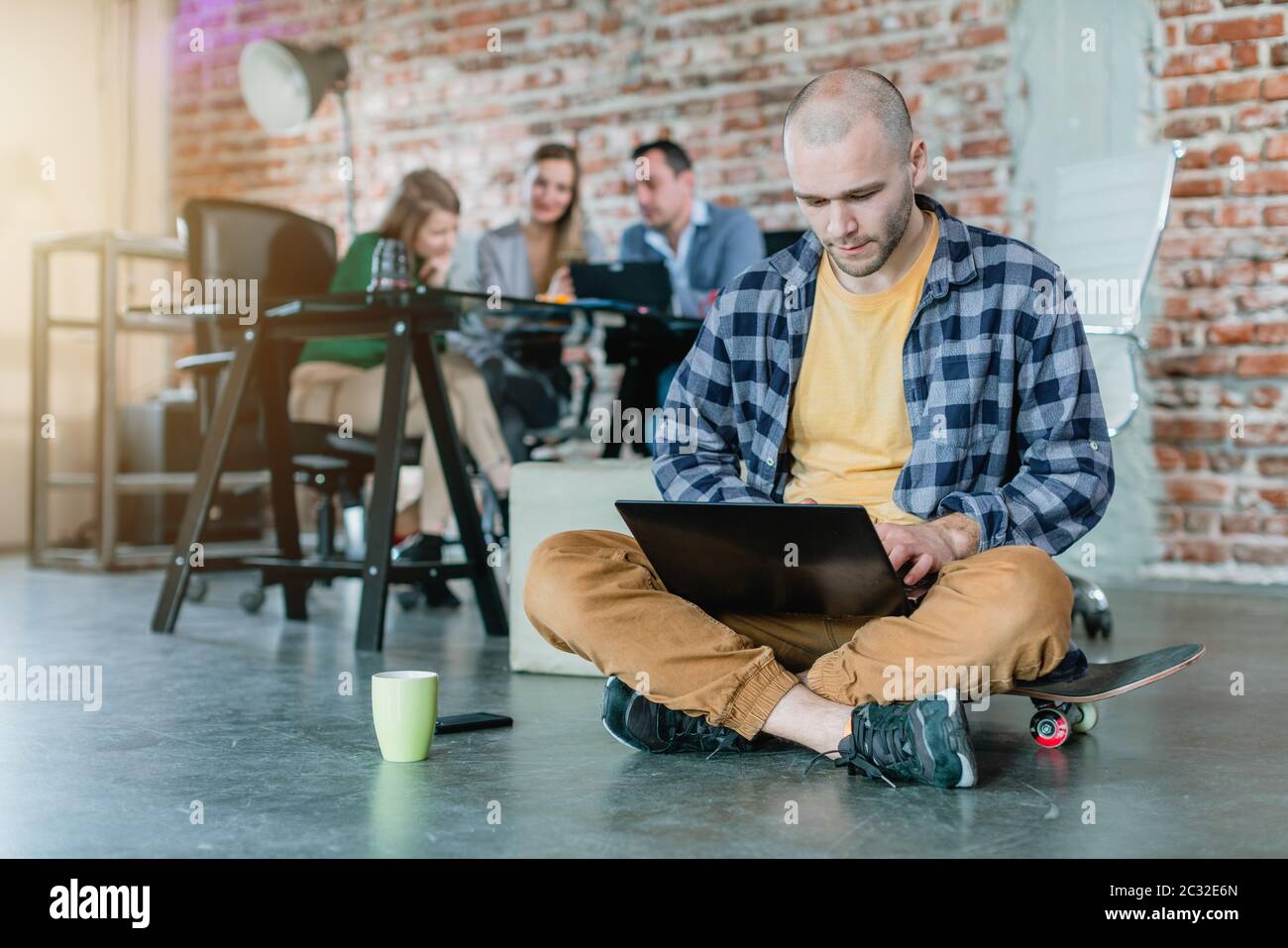 Hip worker in a startup coding with laptop sitting on skateboard with ...