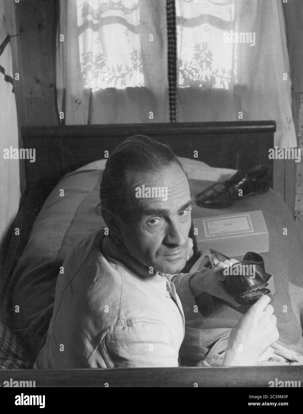 A man sits on his bed, holding a broken shoe in his hand. The photo is ...